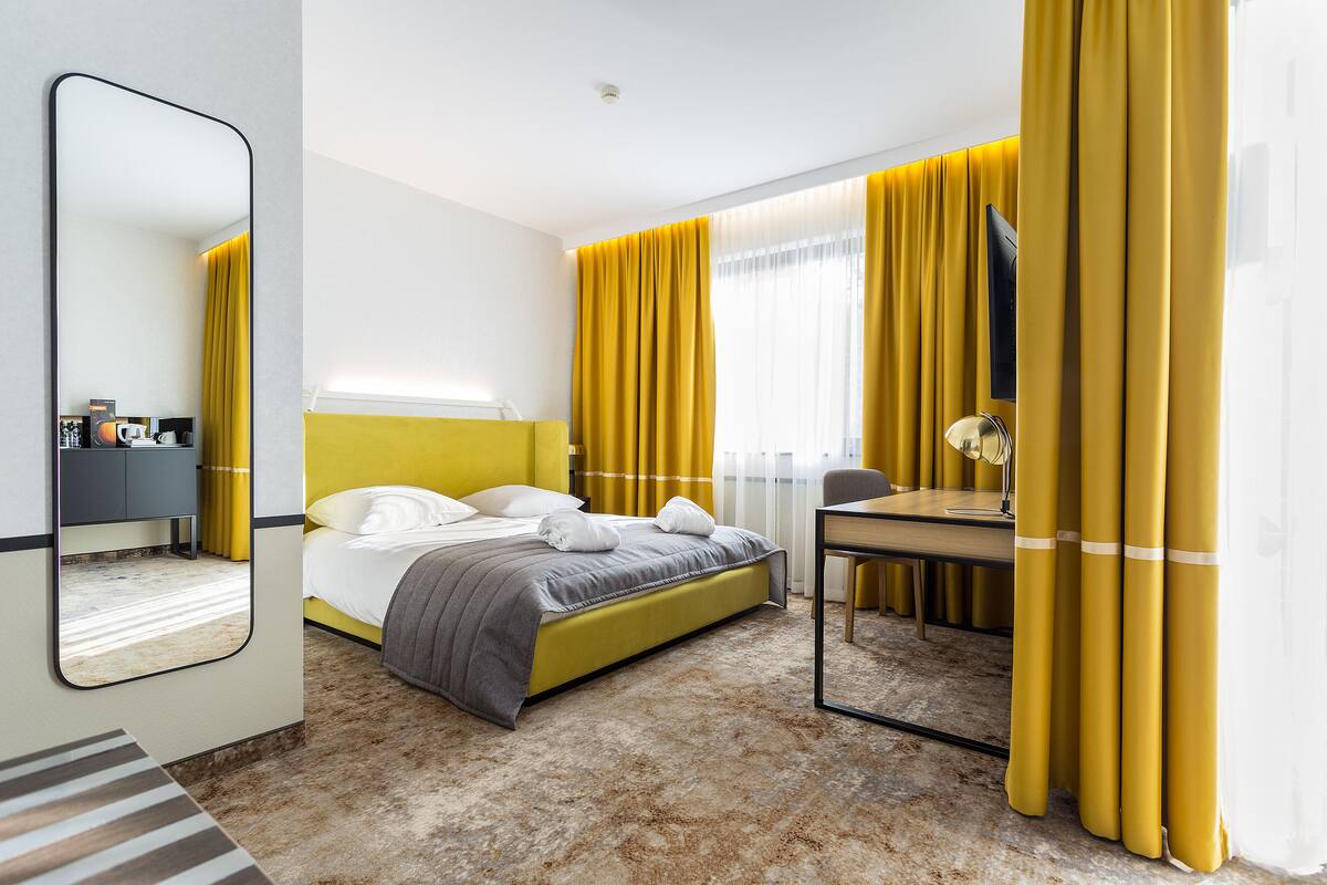 Quarto de hotel com cama, escrivaninha, cadeira, espelho e cortinas.