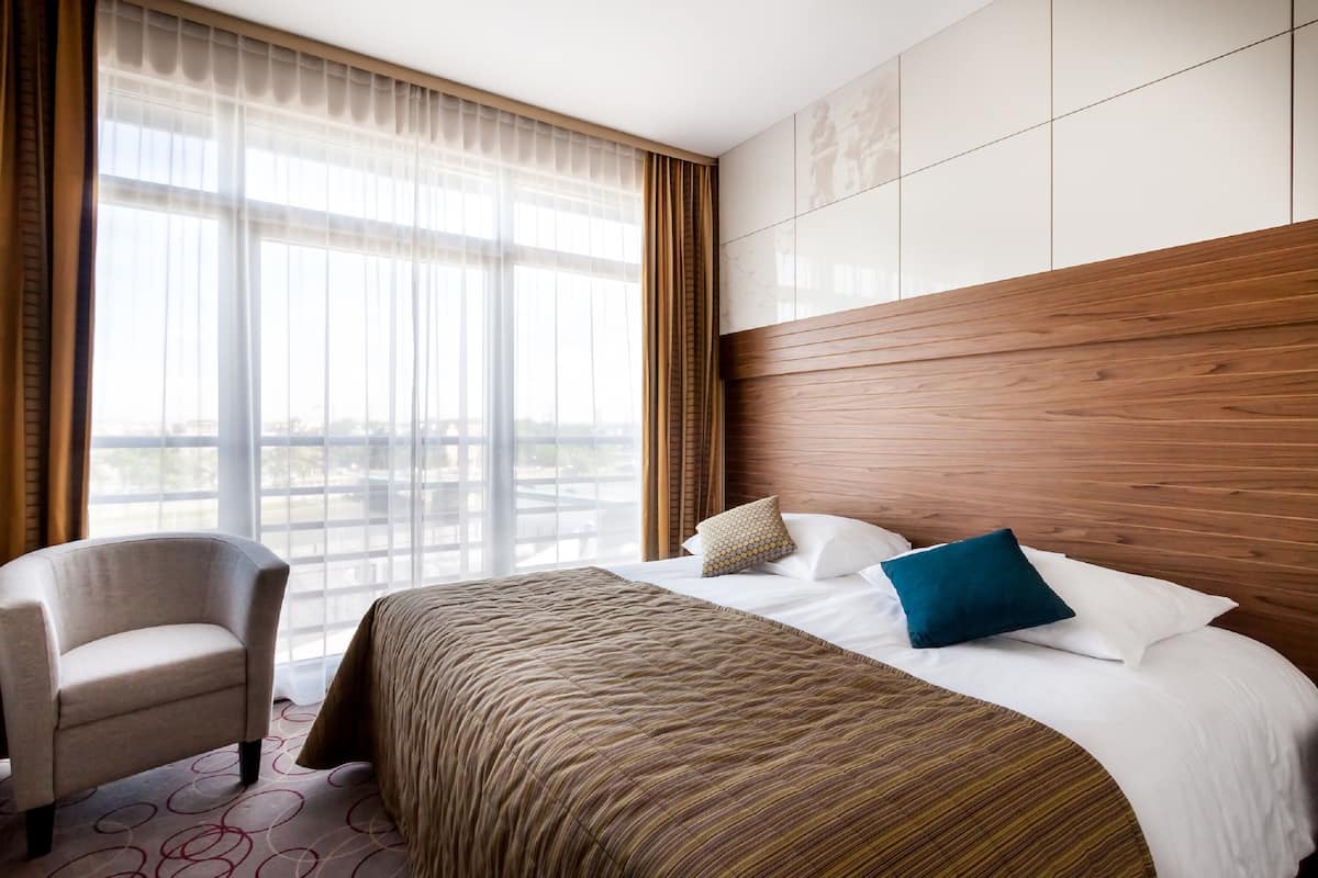 Quarto de hotel com uma grande janela, cama com roupa de cama branca, cabeceira de madeira e uma cadeira.