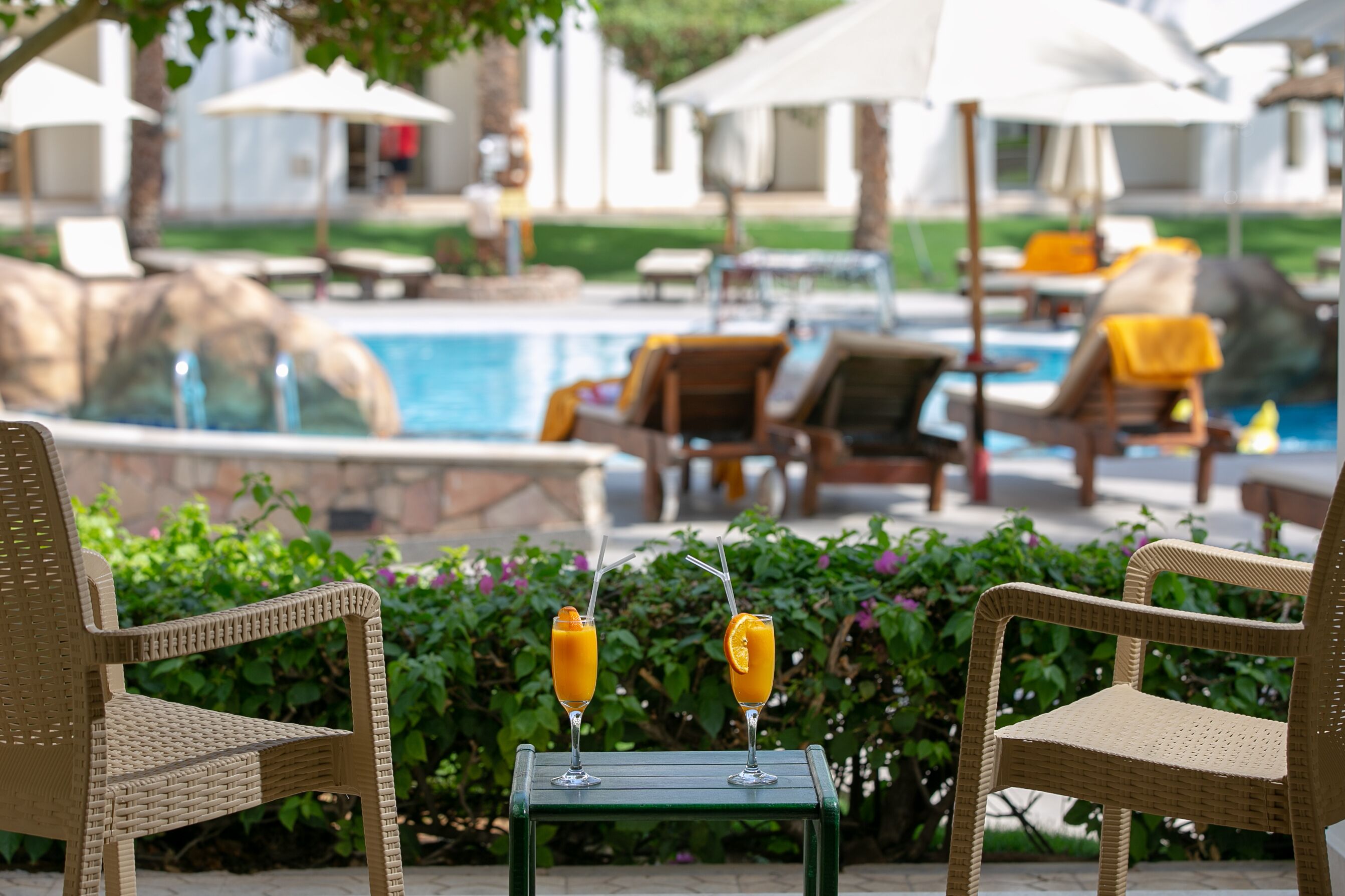 Uma área à beira da piscina com cadeiras de vime, uma mesa pequena com duas bebidas e guarda-sóis ao fundo.