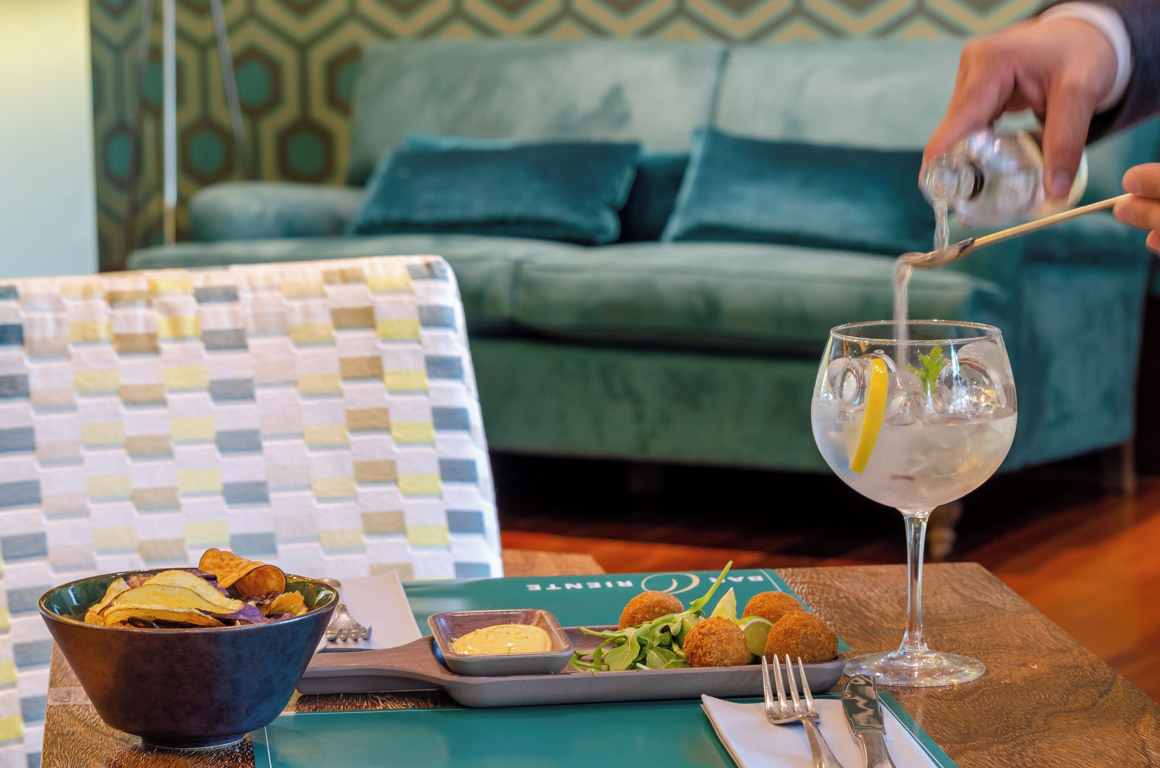 Uma mesa com uma saladeira, um prato de croquetes, um copo de bebida gelada com uma fatia de limão e um pequeno recipiente com molho.