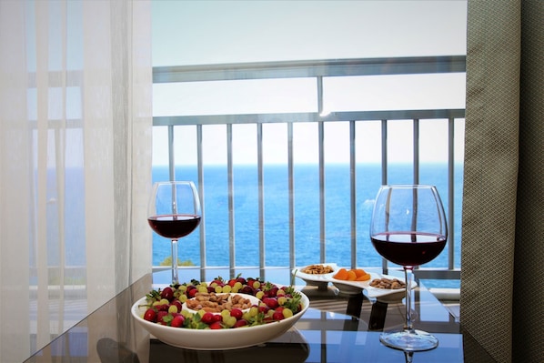 Uma mesa com uma tigela de frutas, dois copos de vinho tinto e um prato de nozes, junto a uma janela com vista para o mar.