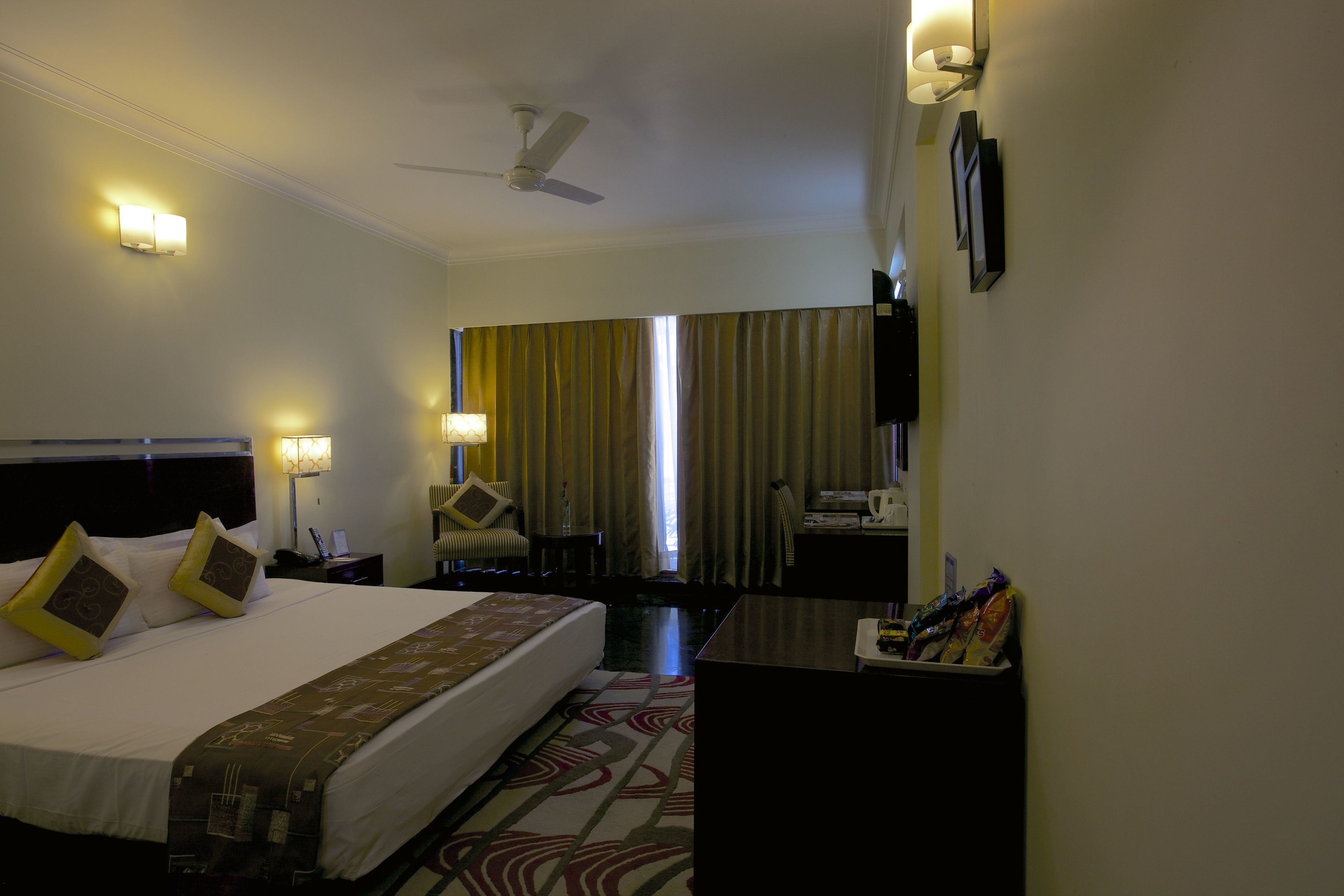 Quarto de hotel com cama, abajures, escrivaninha, cadeira, ventilador de teto e janela com cortinas.