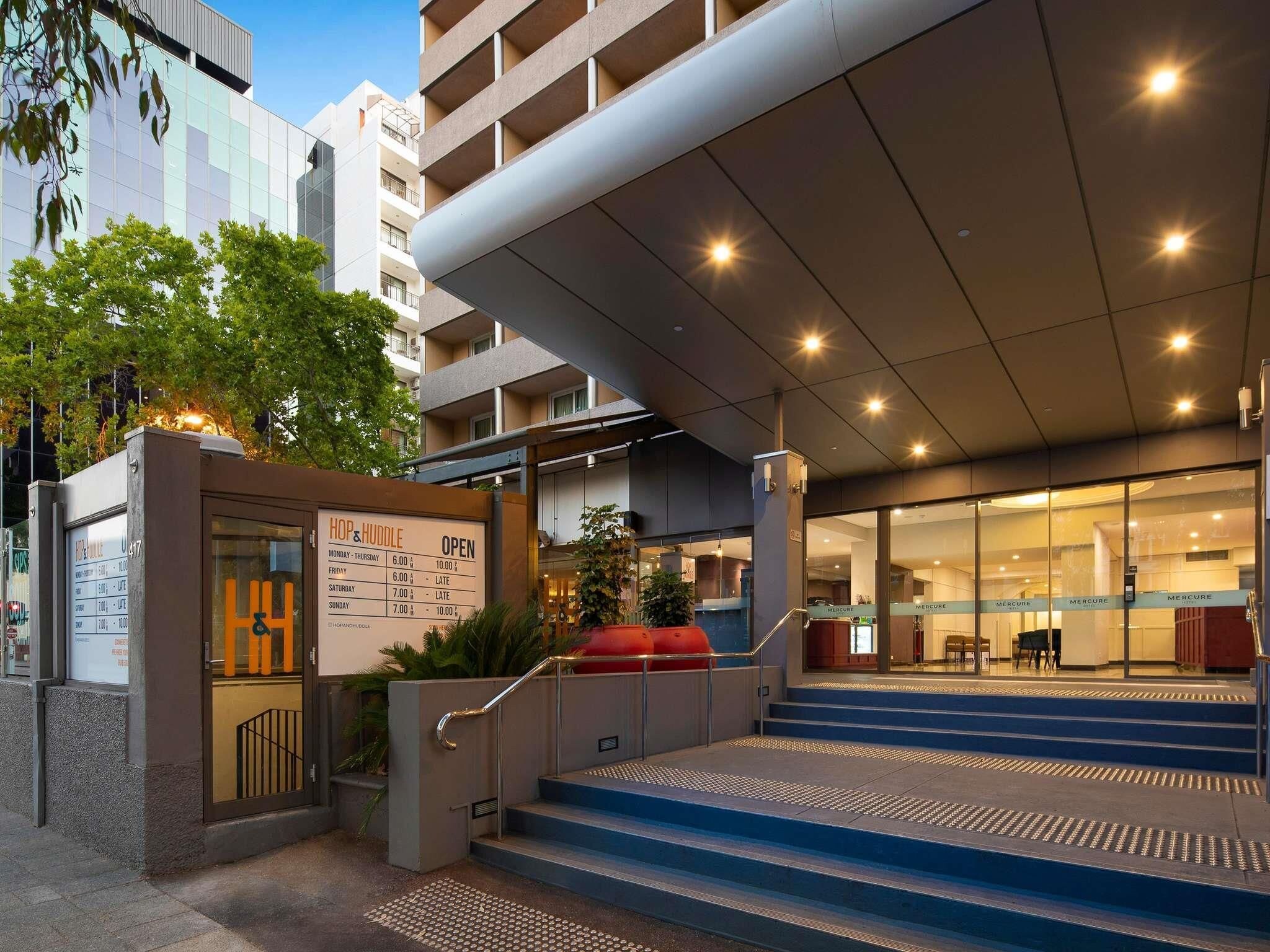 Entrada de hotel com fachada de vidro, área de estar externa e um quadro de menu visível para restaurante.