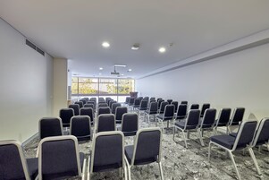 Uma sala de conferências com fileiras de cadeiras, um projetor e uma janela grande com vista para árvores.