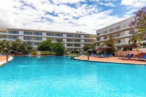 Piscina com espreguiçadeiras e guarda-sóis, rodeada por um edifício com várias varandas.