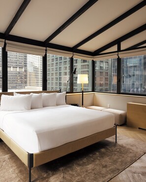 Quarto de hotel moderno com uma cama grande, mesa de cabeceira, luminária e vista da cidade através de amplas janelas.