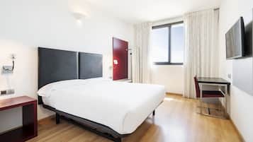 Quarto de hotel com cama, escrivaninha, cadeira, televisão e janela com cortinas.