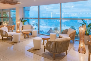 Uma área de estar moderna com móveis de vime, almofadas brancas e grandes janelas com vista para o mar.