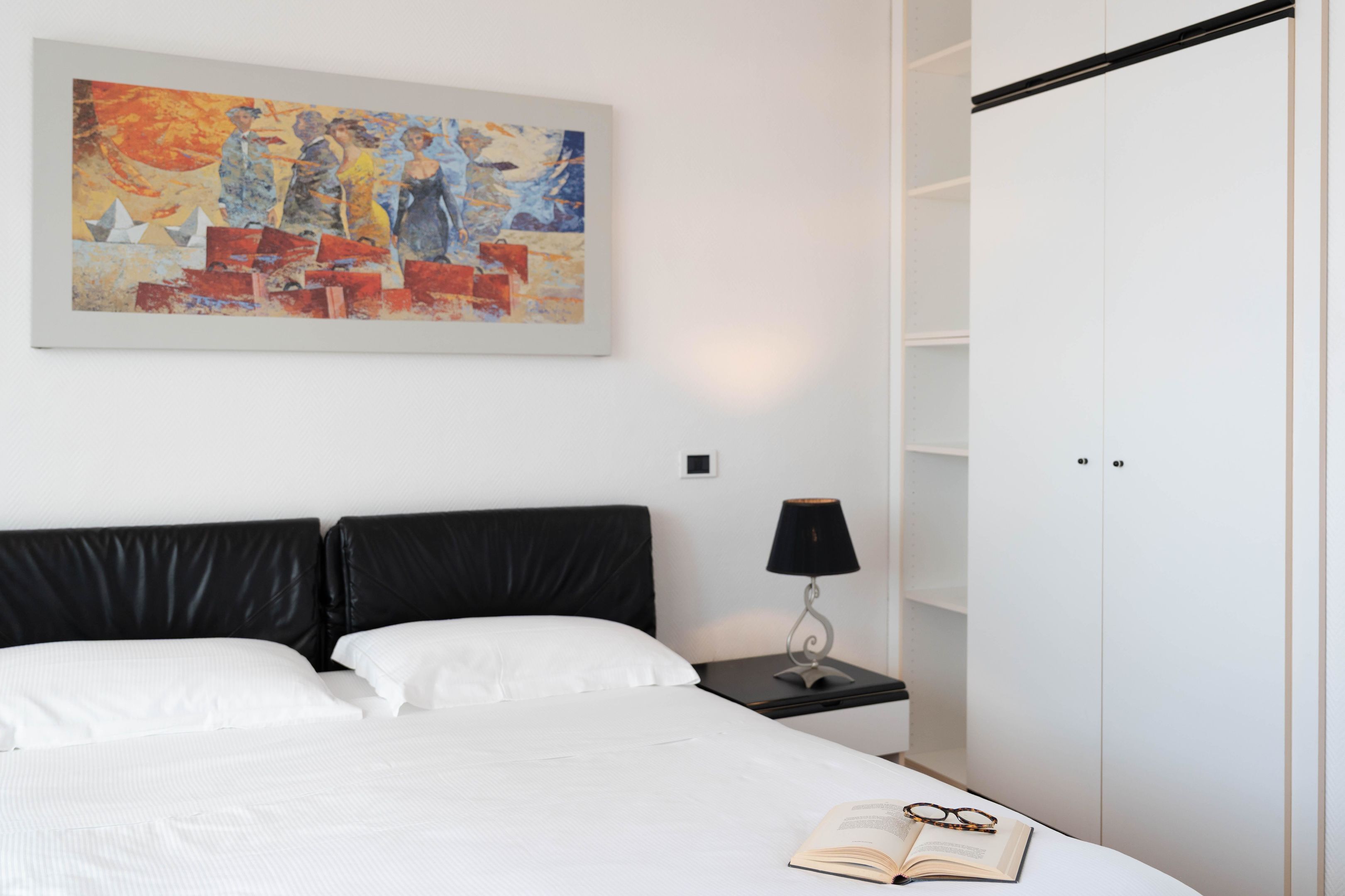 Quarto de hotel com uma cama, mesa de cabeceira, luminária, estante e um quadro na parede.