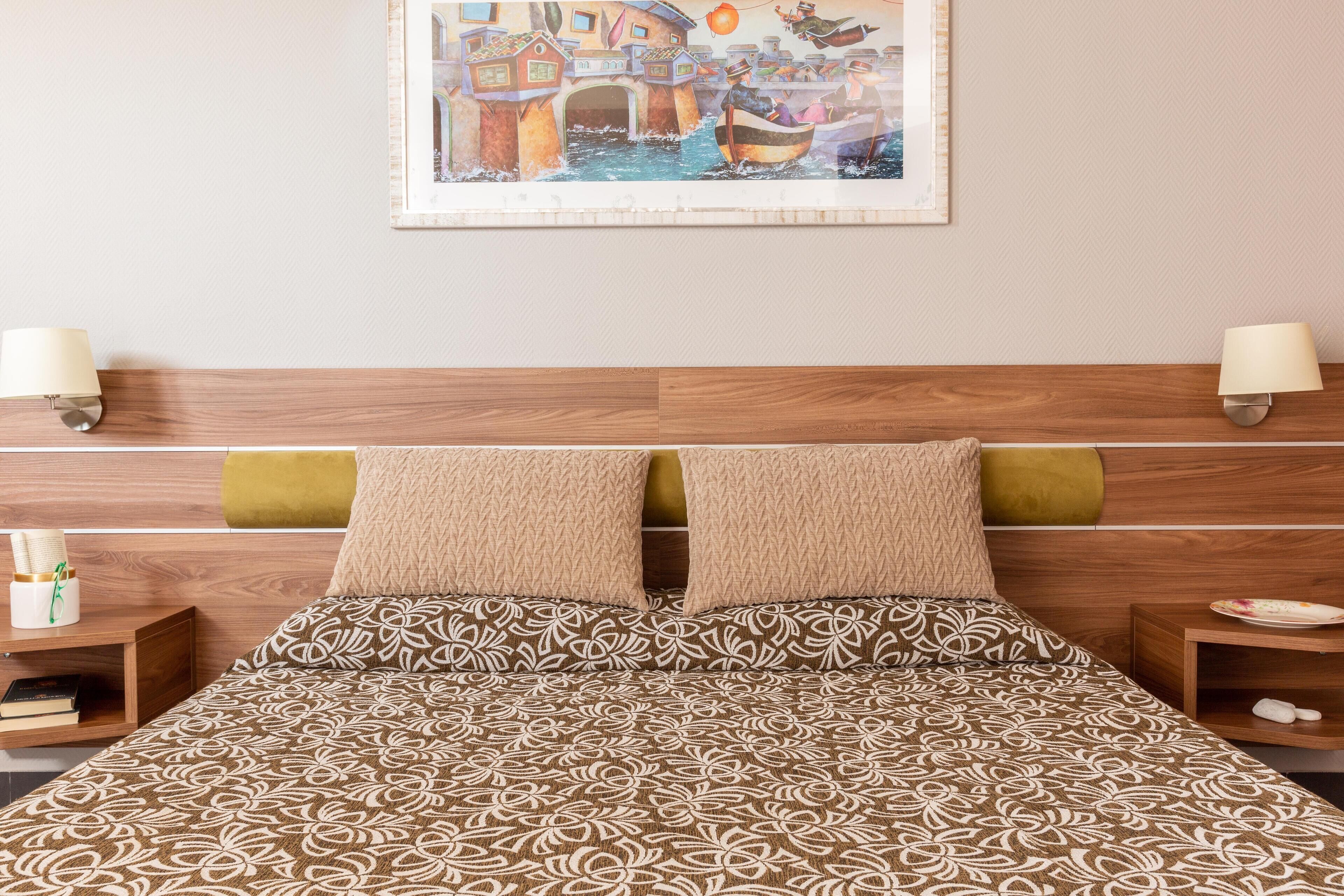 Cama bem arrumada com roupa de cama estampada, cabeceira de madeira e duas mesas de cabeceira com abajures.