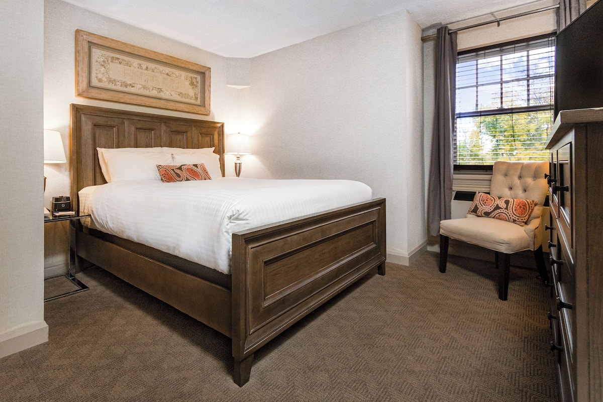 Quarto com cama de madeira, uma cadeira, mesa de cabeceira e janela com persianas.