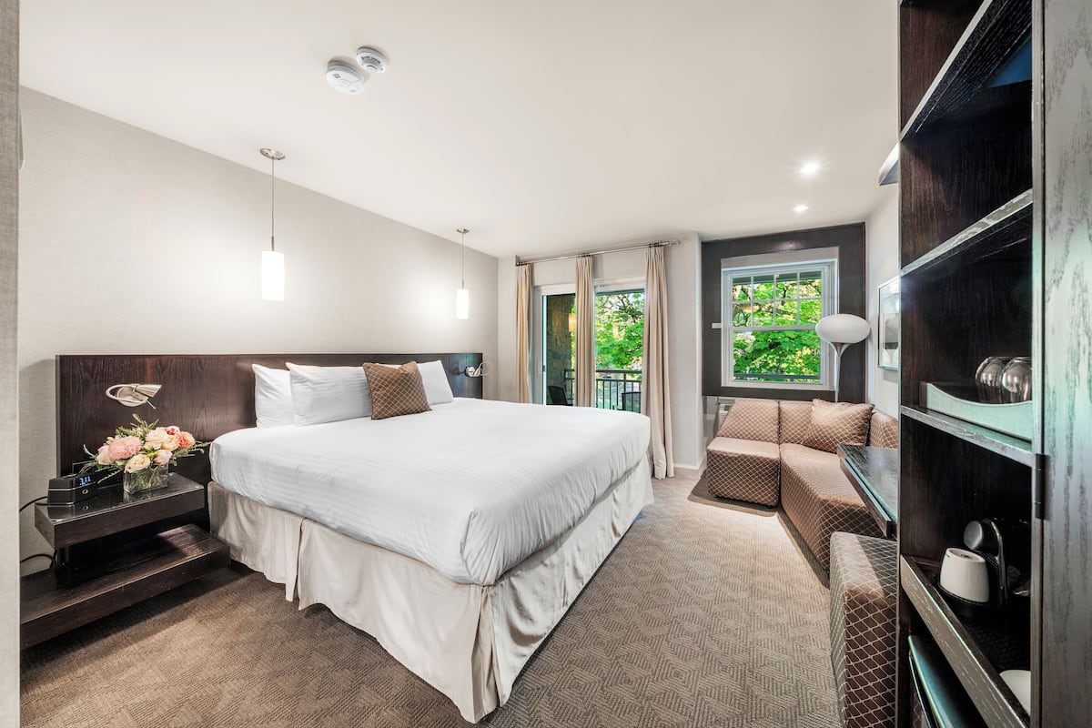 Quarto de hotel moderno com uma cama grande, uma mesa de cabeceira, um espaço de estar e vista para o verde lá fora.