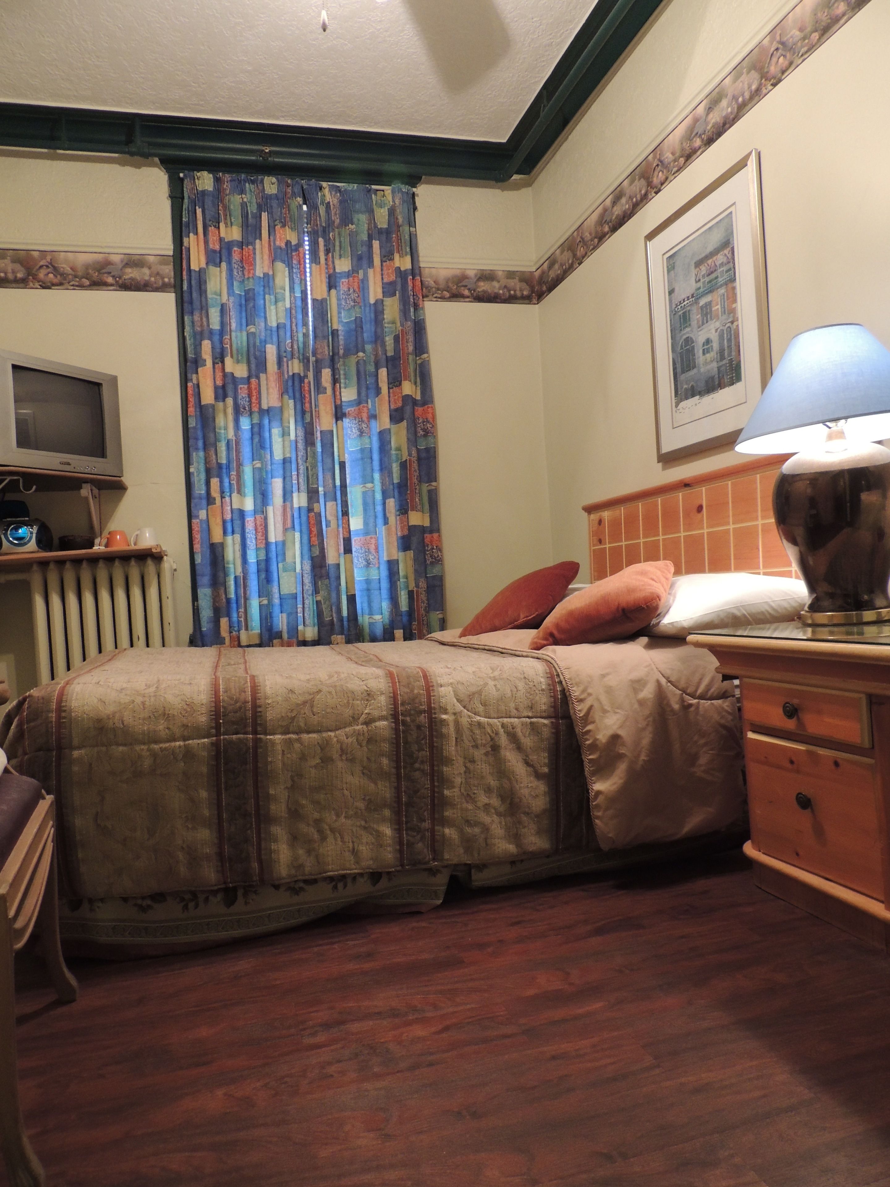 Quarto com cama, penteadeira, luminária, televisão em um móvel e cortina.