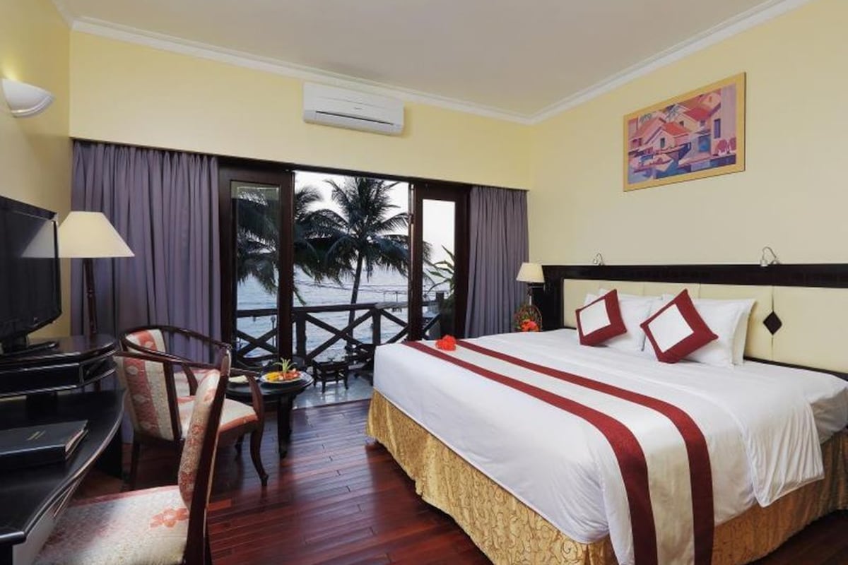 Quarto de hotel com cama, escrivaninha, cadeira e vista para palmeiras através de uma varanda.