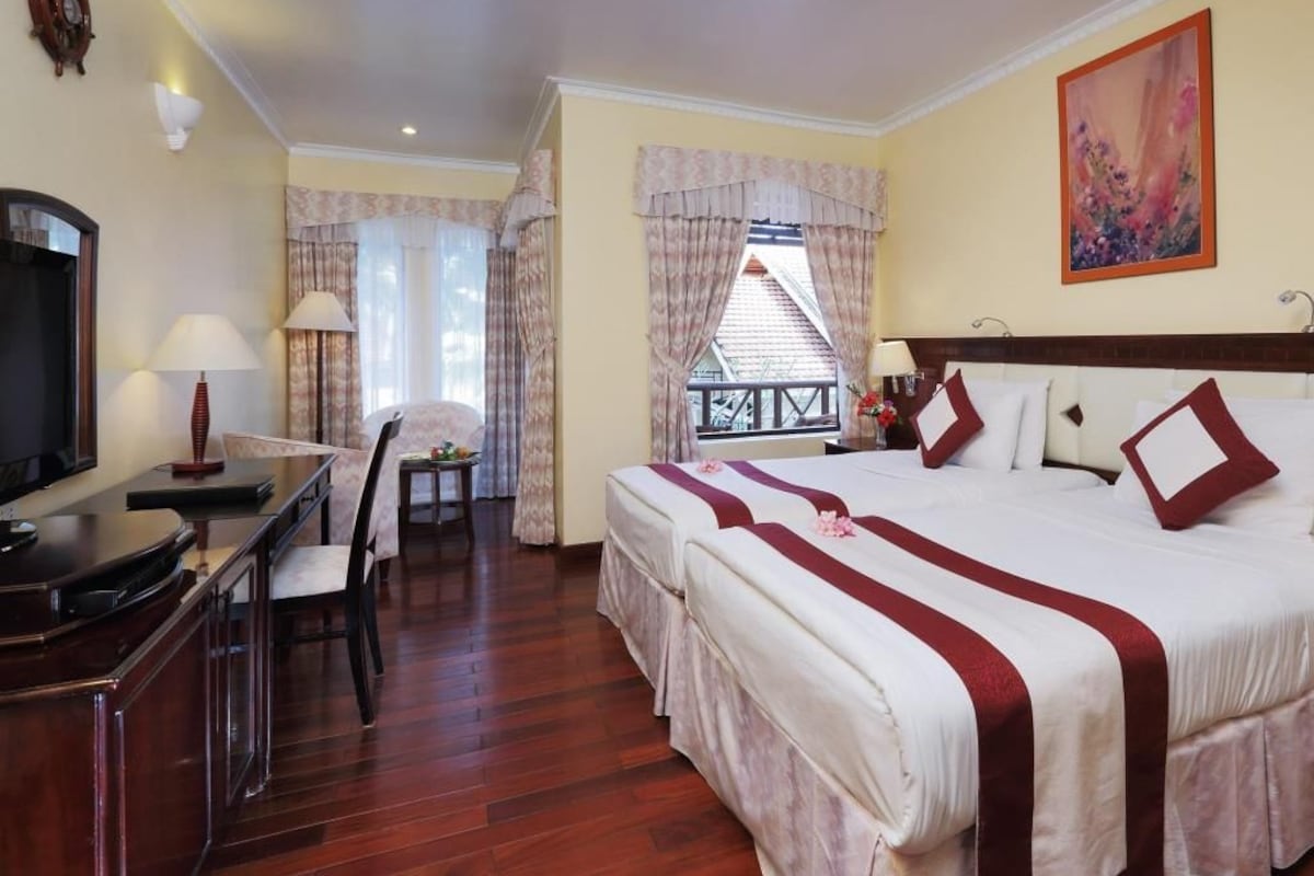 Quarto de hotel com uma cama grande, uma mesa com cadeira, televisão e uma varanda com cortinas.