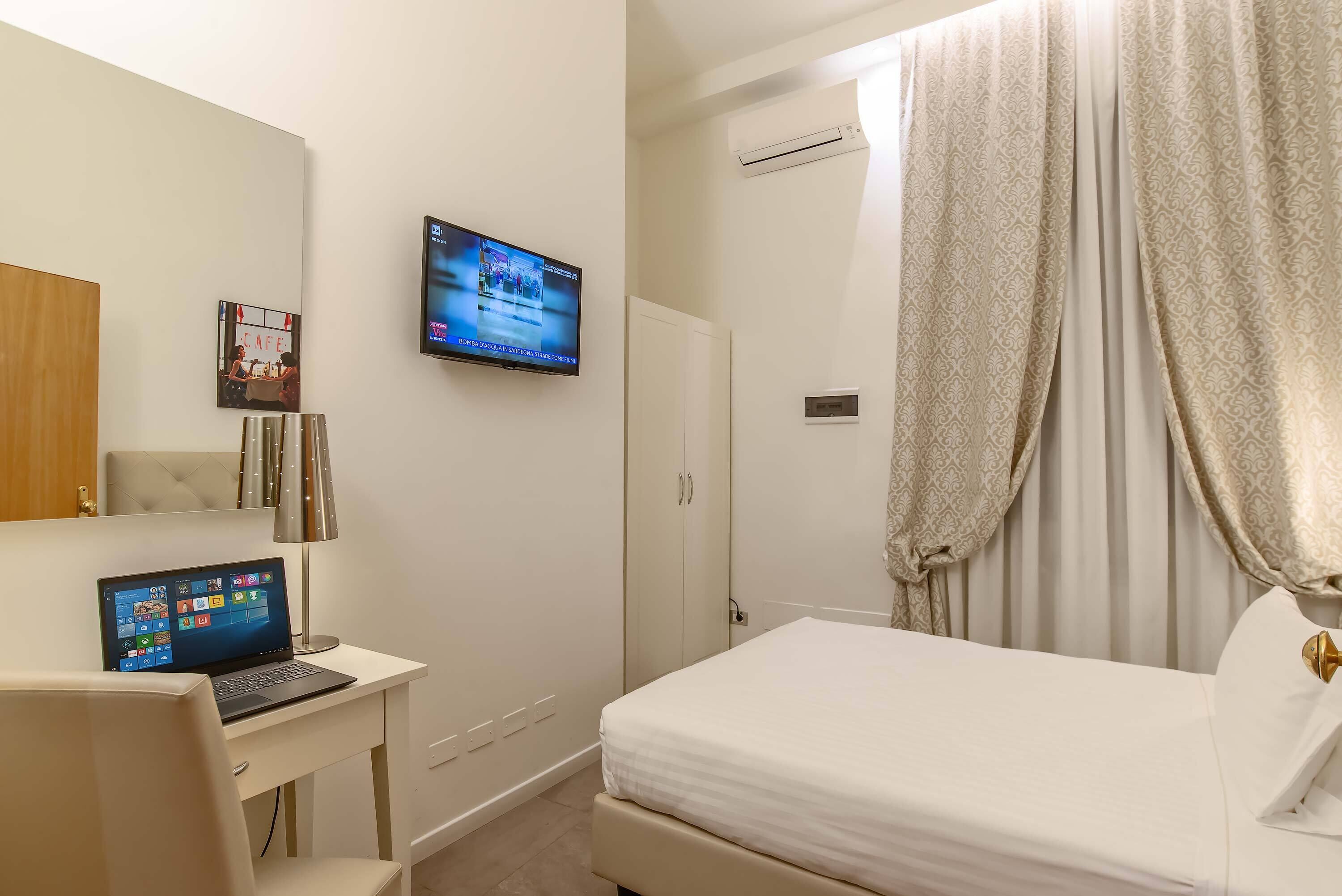 Quarto de hotel com cama, secretária com laptop, televisão na parede e guarda-roupa.