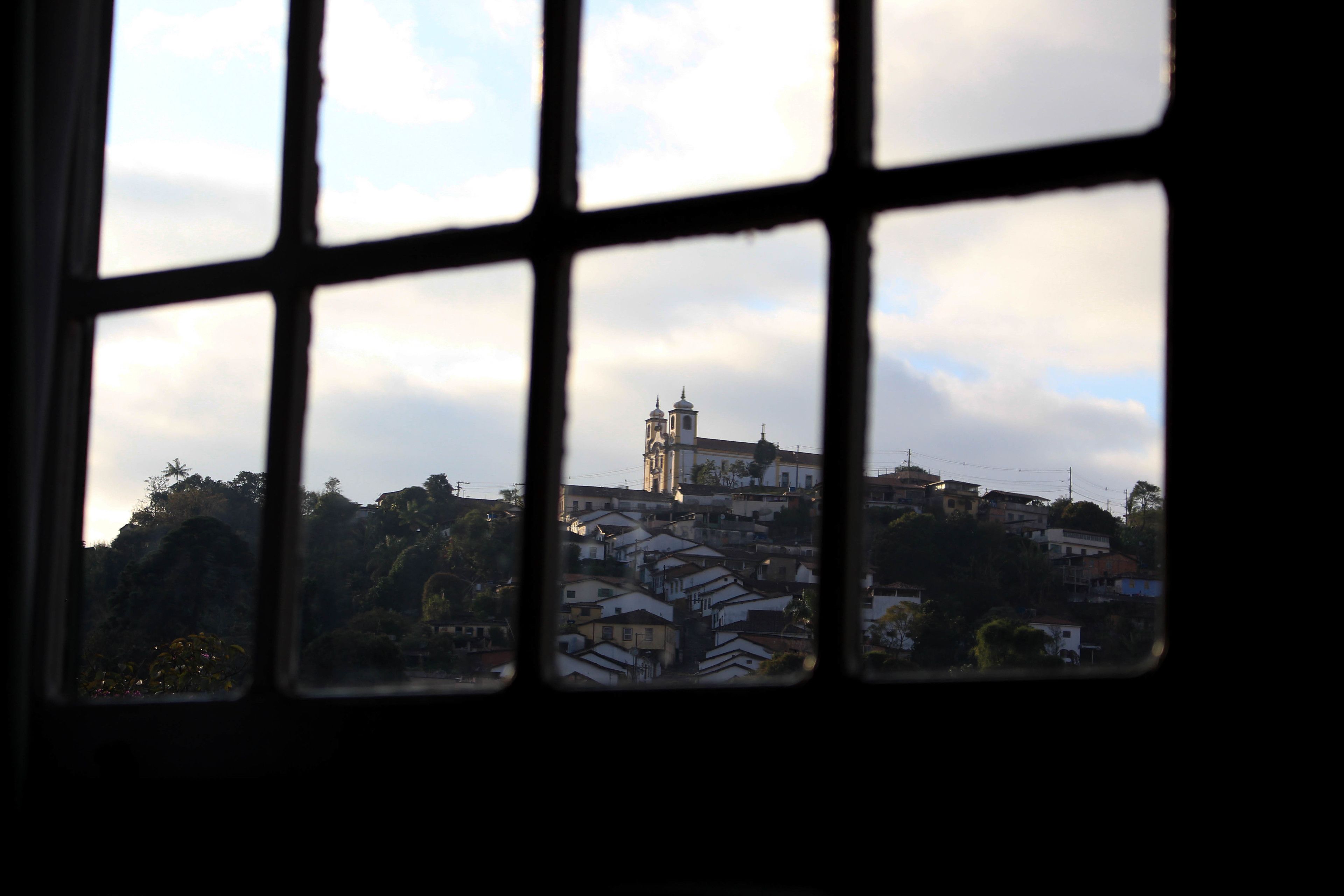 Vista pela janela, com padrão quadriculado, mostrando uma cidade com edifícios e uma torre de igreja imponente.