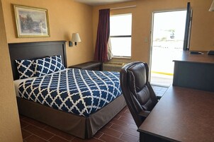 Quarto de hotel com cama, escrivaninha, cadeira, abajur e janela com cortinas.