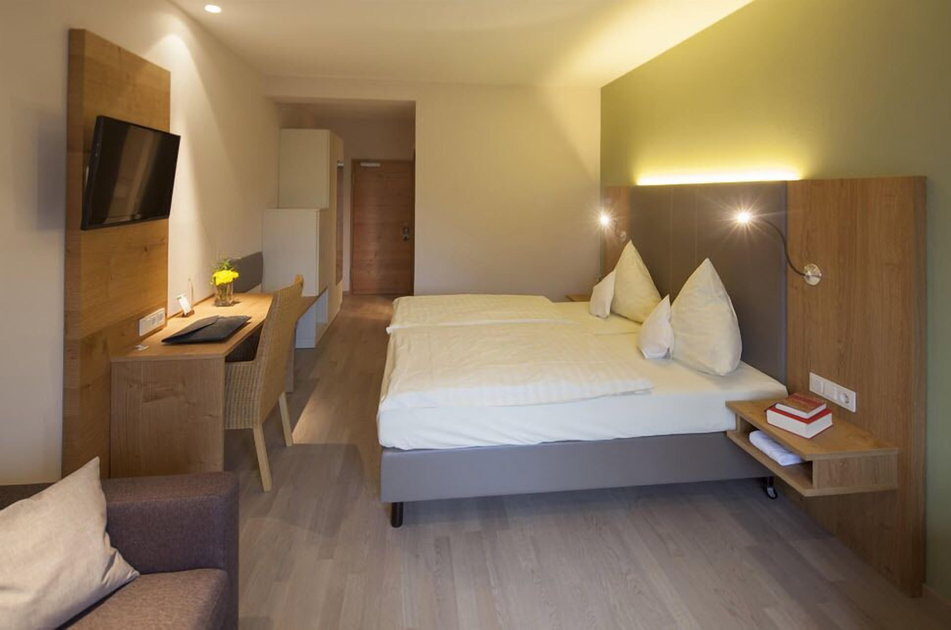 Quarto de hotel com uma cama, uma escrivaninha com cadeira, uma televisão na parede e uma mesa de cabeceira com um livro.