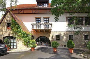 Um edifício de pedra com design tradicional, apresentando uma entrada em arco central, uma varanda com grades e plantas em vasos.