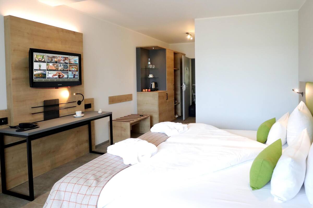 Quarto de hotel com cama, escrivaninha, televisão fixada na parede e pequena área de cozinha.