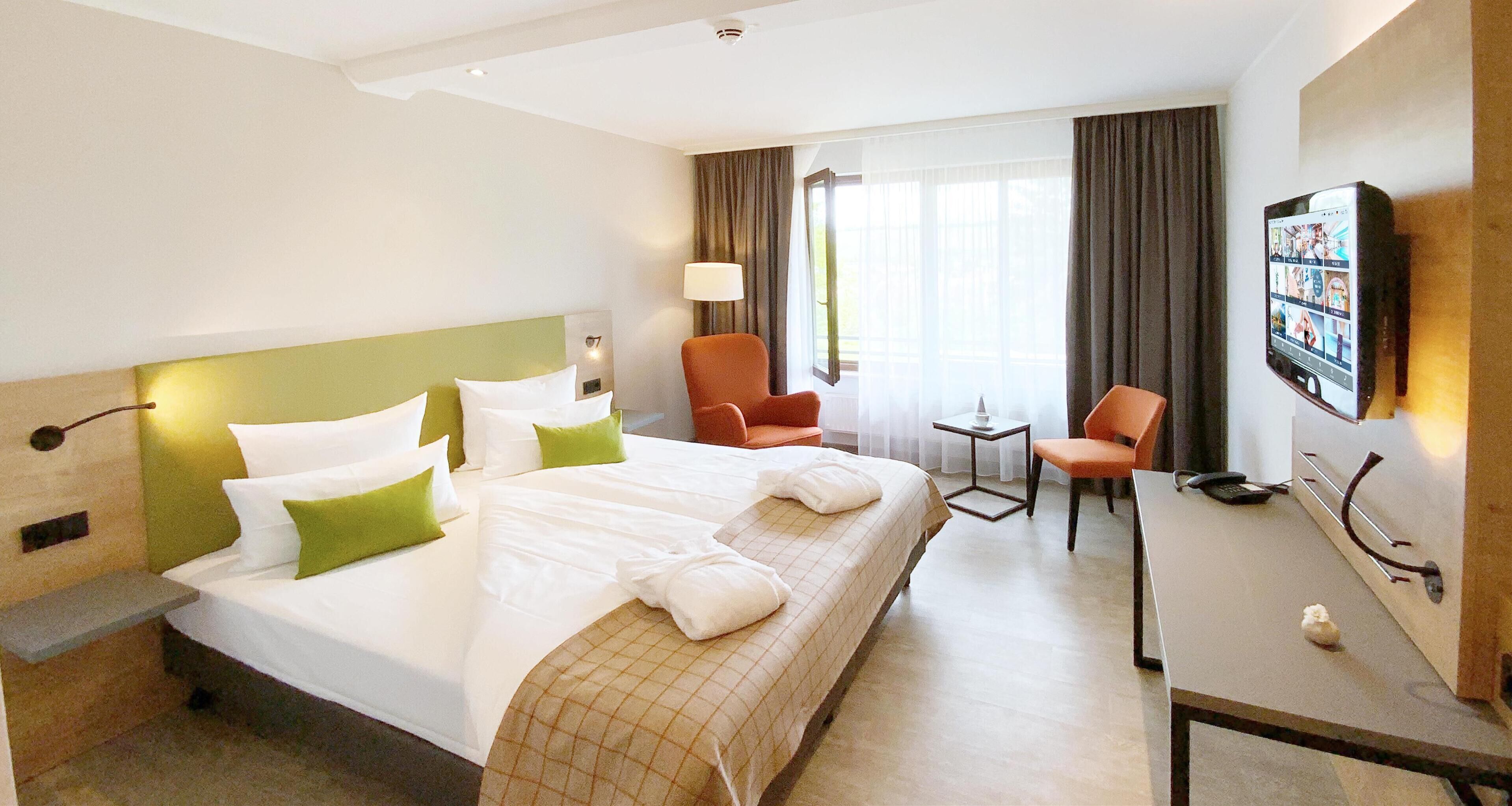 Quarto de hotel com uma cama grande, duas cadeiras, uma escrivaninha, televisão e janela com cortinas.
