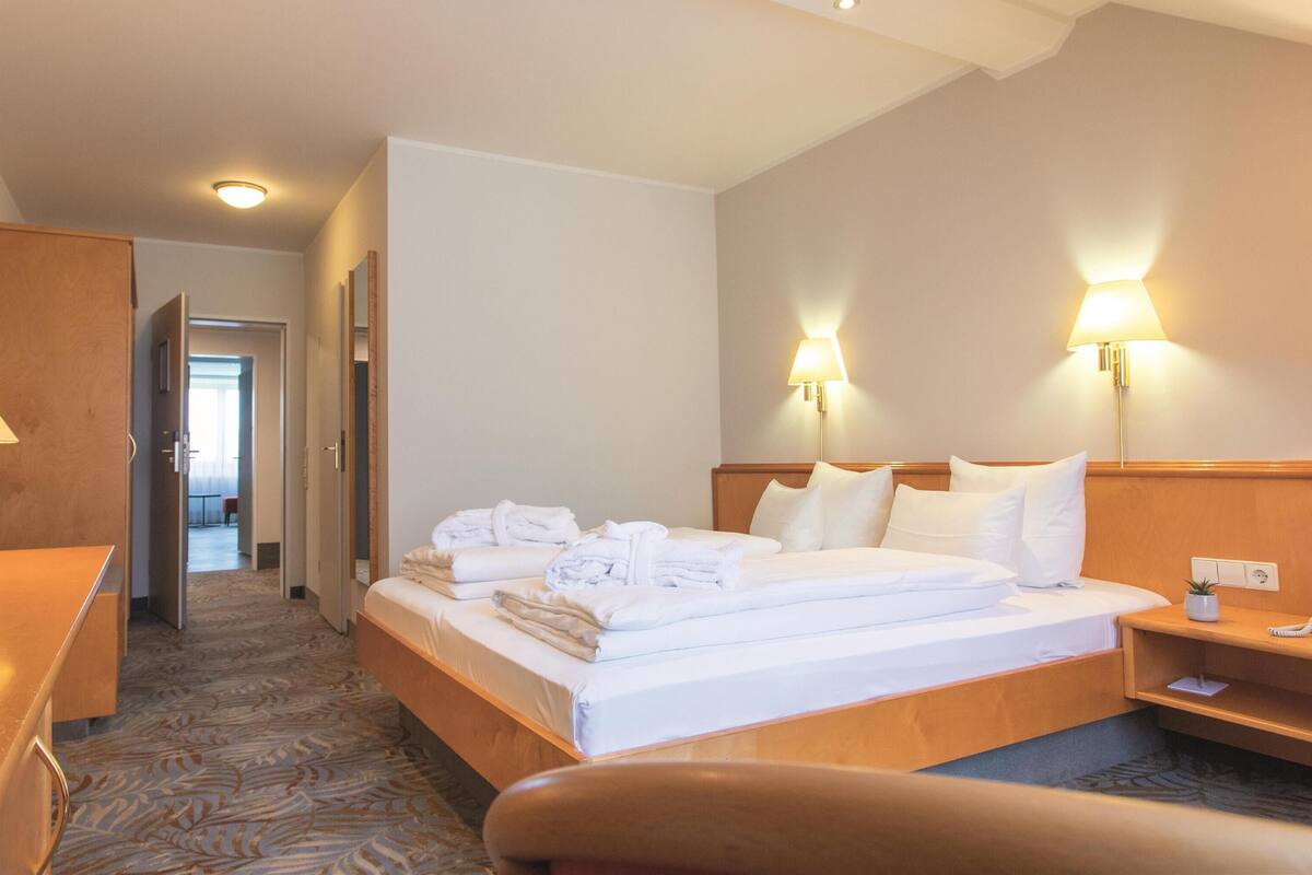 Quarto de hotel com cama bem arrumada, luminárias de cabeceira, escrivaninha e uma cadeira.