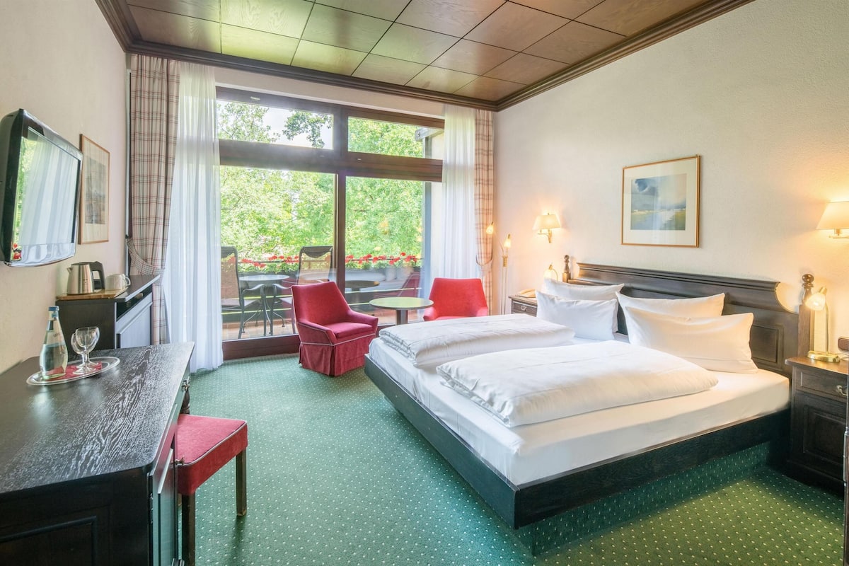 Quarto de hotel com uma cama grande, uma escrivaninha com cadeira, uma mesinha e uma janela com cortinas.