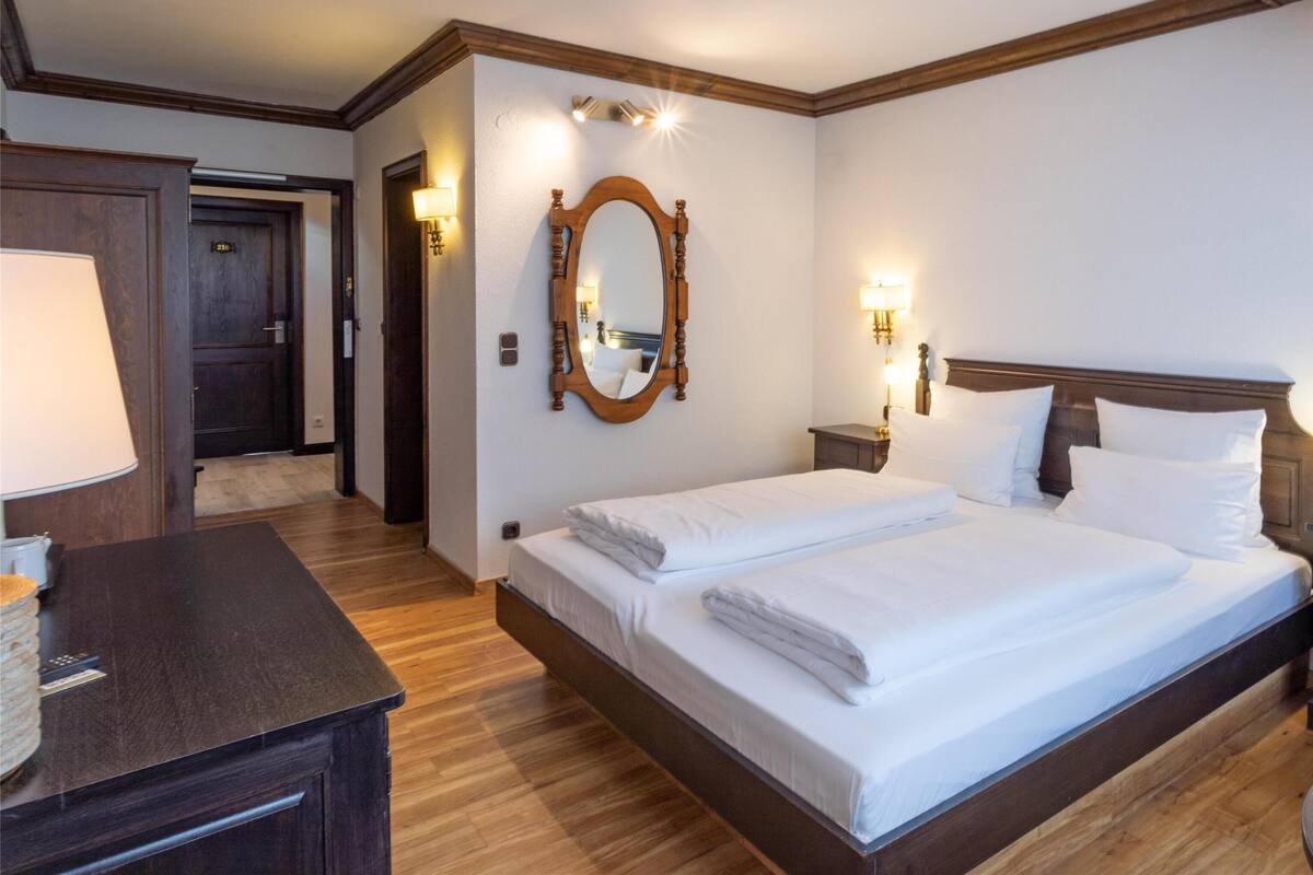 Quarto de hotel com uma cama grande, cabeceira de madeira, espelho, abajures, escrivaninha com luminária e uma cadeira.