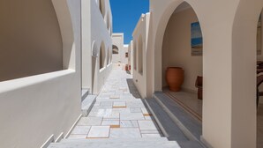 Um corredor estreito, com paredes brancas e chão de azulejos, e aberturas em arco, que leva a um edifício sob um céu azul claro.