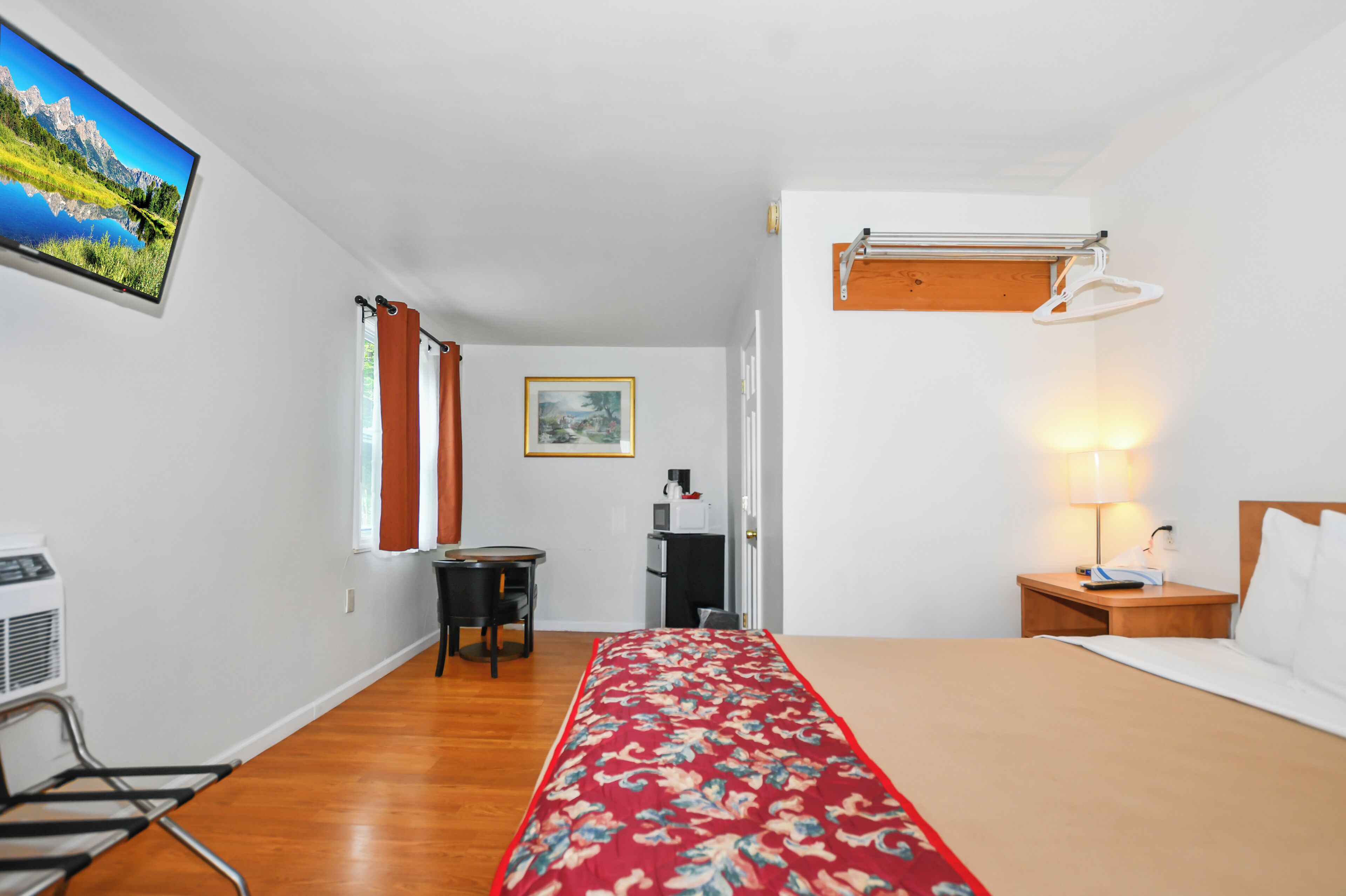 Quarto de hotel com cama, escrivaninha, cadeira, televisão, janela com cortinas, quadro na parede e abajur.