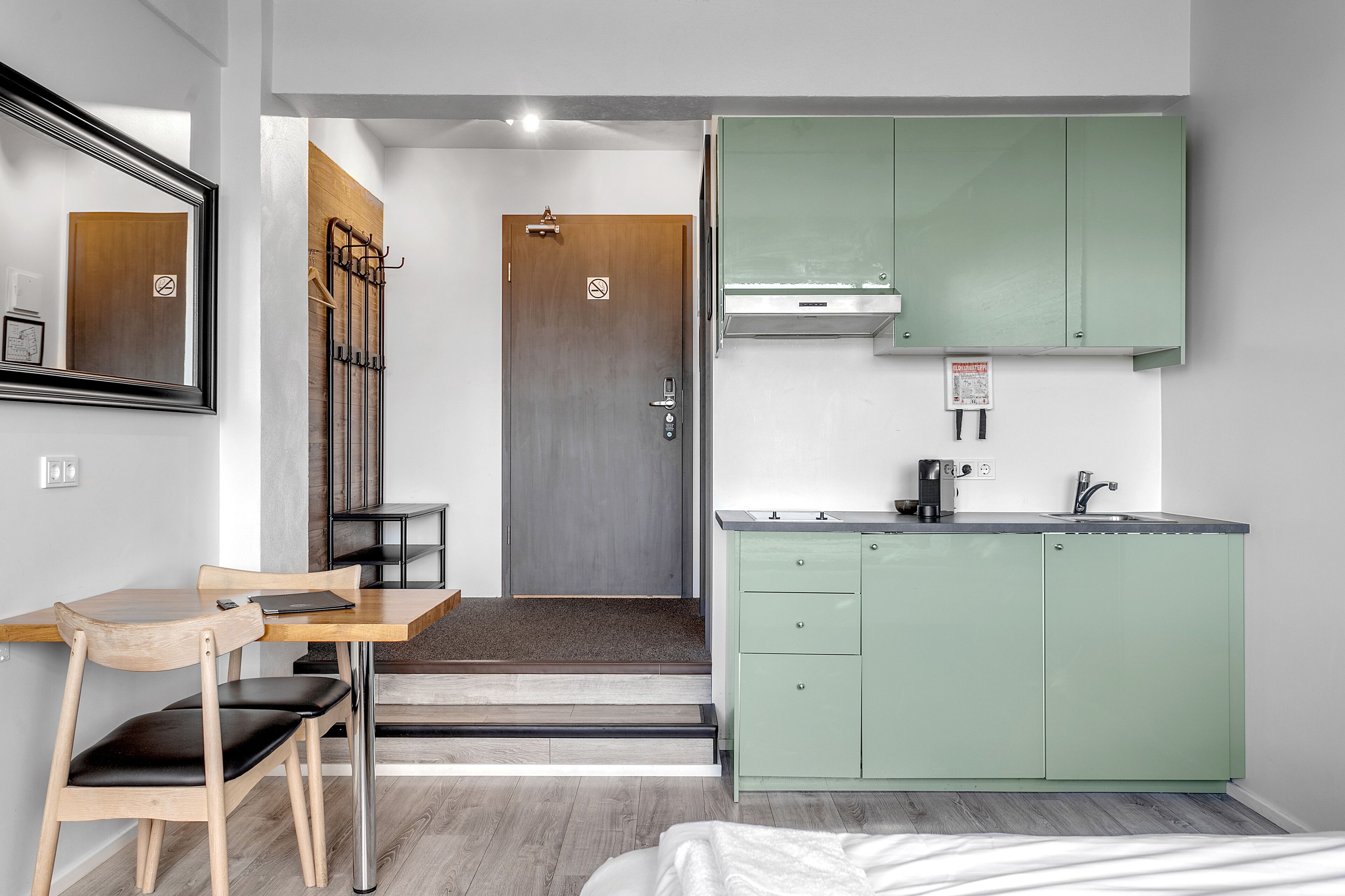 Quarto de hotel pequeno com cozinha compacta, mesa de jantar de madeira com cadeiras, cama e espelho na parede.