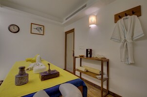 Um quarto com mesa de massagem, toalhas, prateleira de madeira com artigos de higiene pessoal, relógio e quadro na parede.