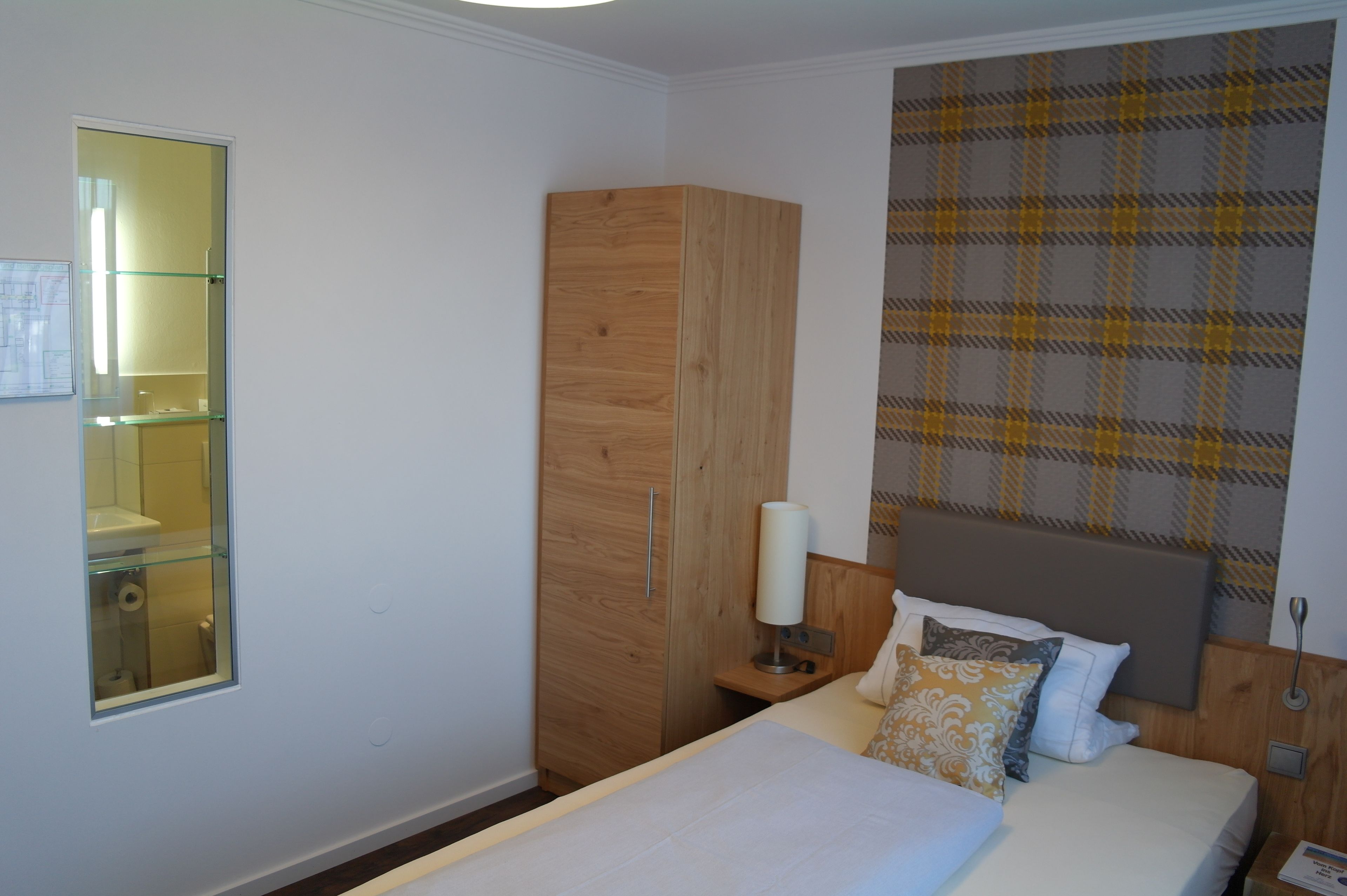 Quarto de hotel com cama, guarda-roupa de madeira, mesa de cabeceira com abajur e cabeceira estampada xadrez.