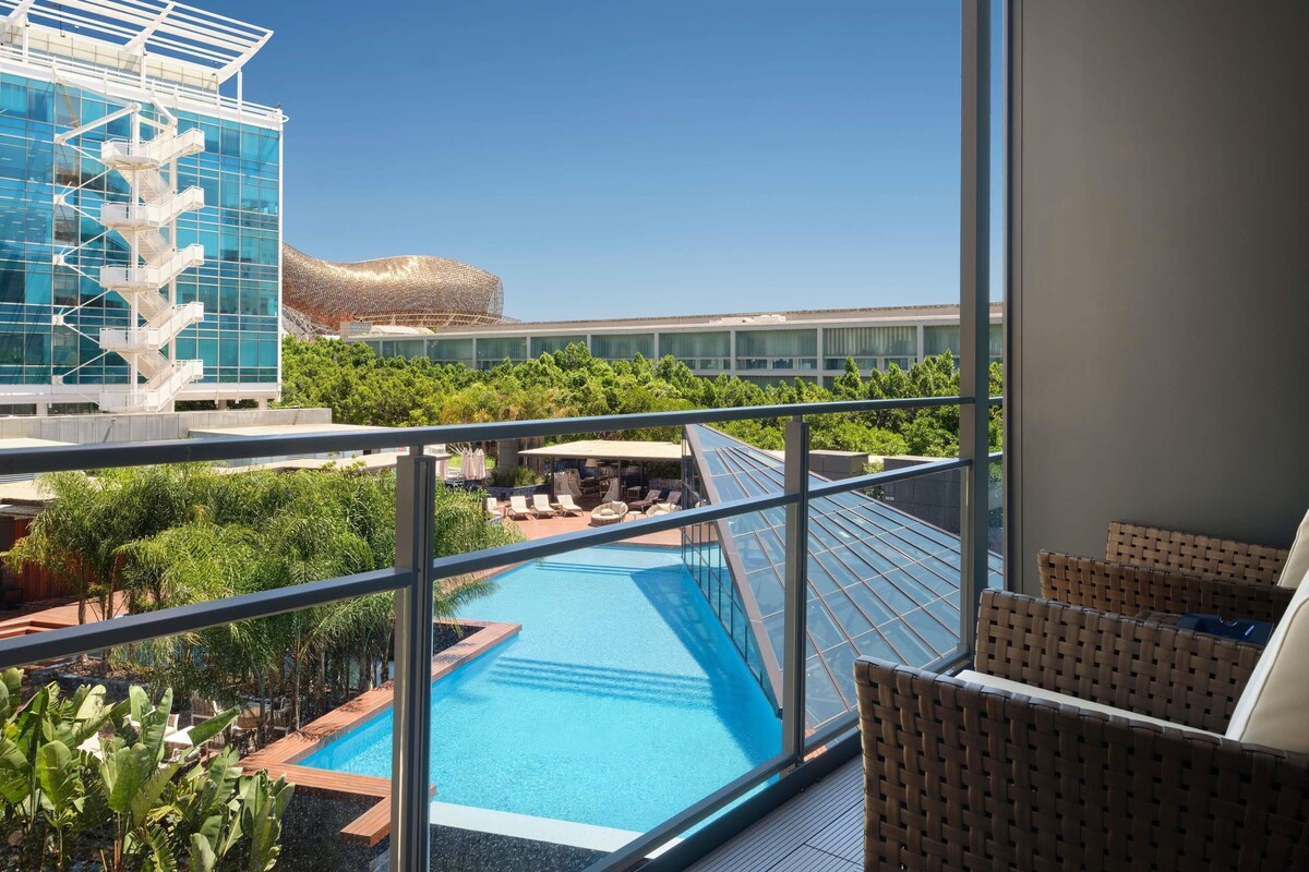 Vista da piscina com guarda-corpo de vidro, cadeiras de descanso e um edifício moderno com estrutura de telhado singular.