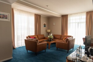 Quarto de hotel com carpete azul, dois sofás de couro marrom, uma mesa de centro de vidro, uma televisão e um quadro na parede.