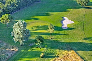 Vista aérea de um campo de golfe com bunkers de areia, árvores e céu claro.
