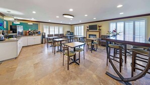 Uma ampla sala de jantar com várias mesas e cadeiras, área de cozinha com geladeira e bancada, e lareira.