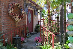 Uma casa de tijolos com porta e escadaria vermelhas, e um jardim com diversas plantas e uma guirlanda decorativa.