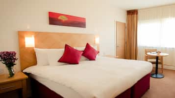 Quarto de hotel com uma cama grande, mesa de cabeceira, um vaso de flores, um quadro na parede e uma janela com cortinas.