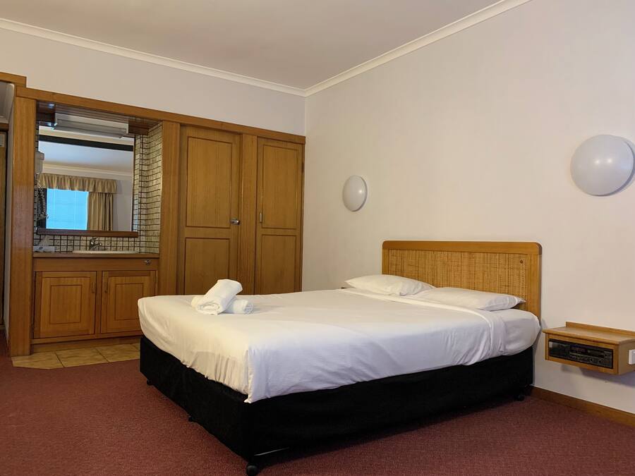 Quarto de hotel com cama, guarda-roupa de madeira, espelho, luz na parede e uma pequena prateleira de madeira.