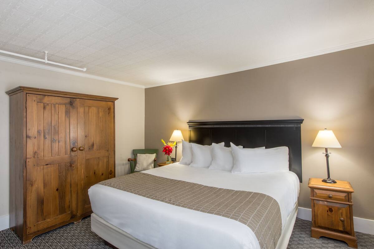 Quarto de hotel com uma cama grande, um guarda-roupa de madeira, duas mesas de cabeceira com luminárias e uma mesinha de cabeceira com abajur.