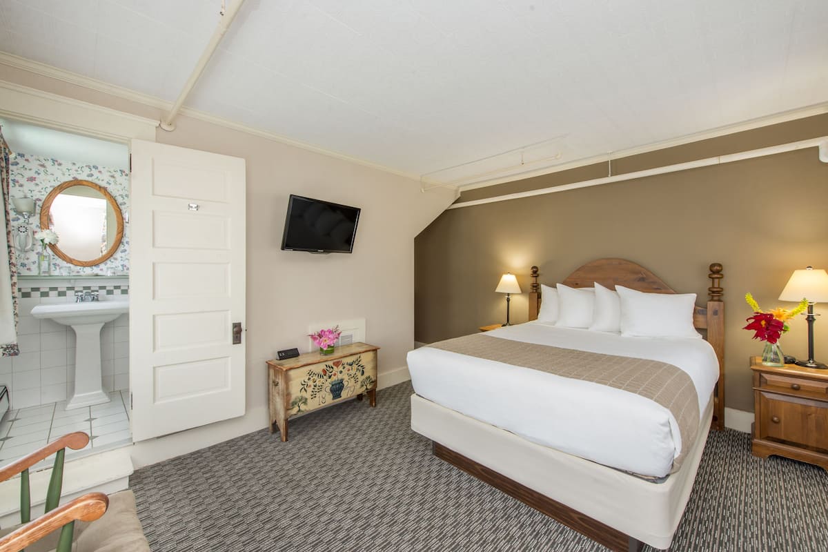 Quarto de hotel com cama, televisão, mesa de cabeceira com abajur, criado-mudo com um vaso de flores e banheiro com pia e espelho.