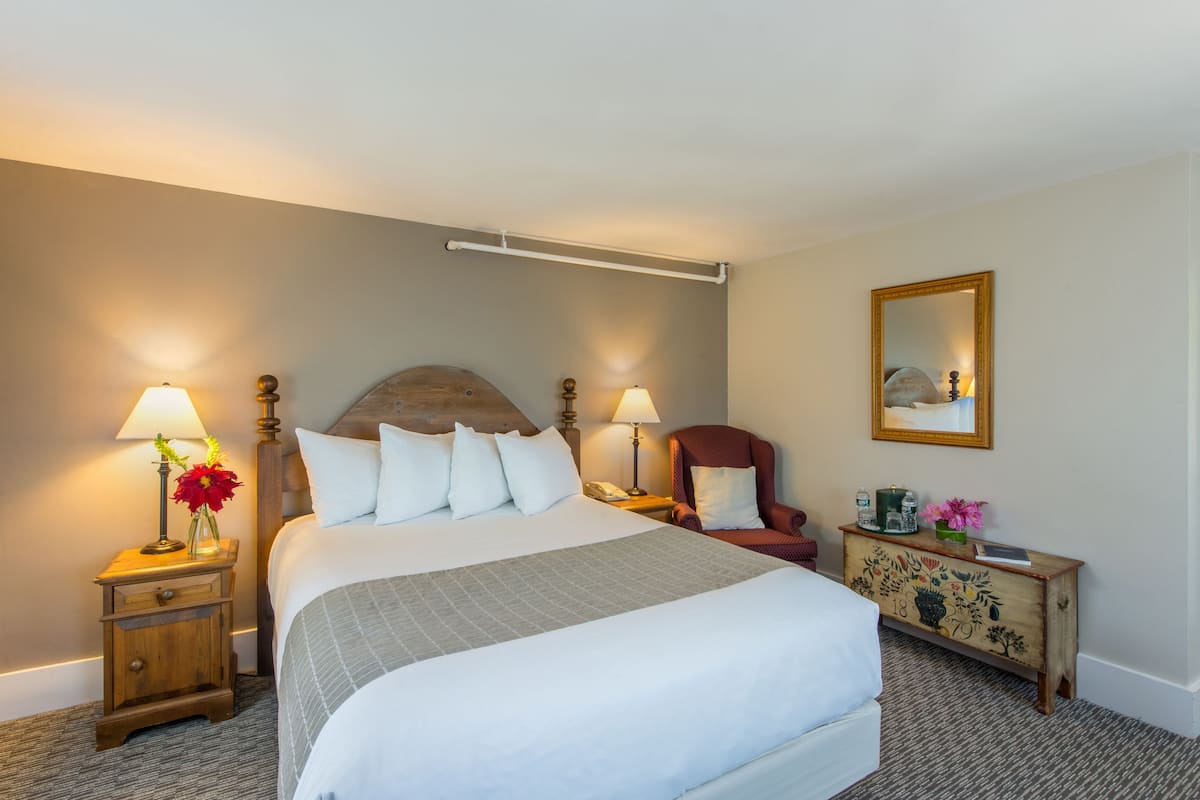 Quarto de hotel com uma cama grande, duas mesas de cabeceira com luminárias, uma cadeira, cômoda e espelho na parede.