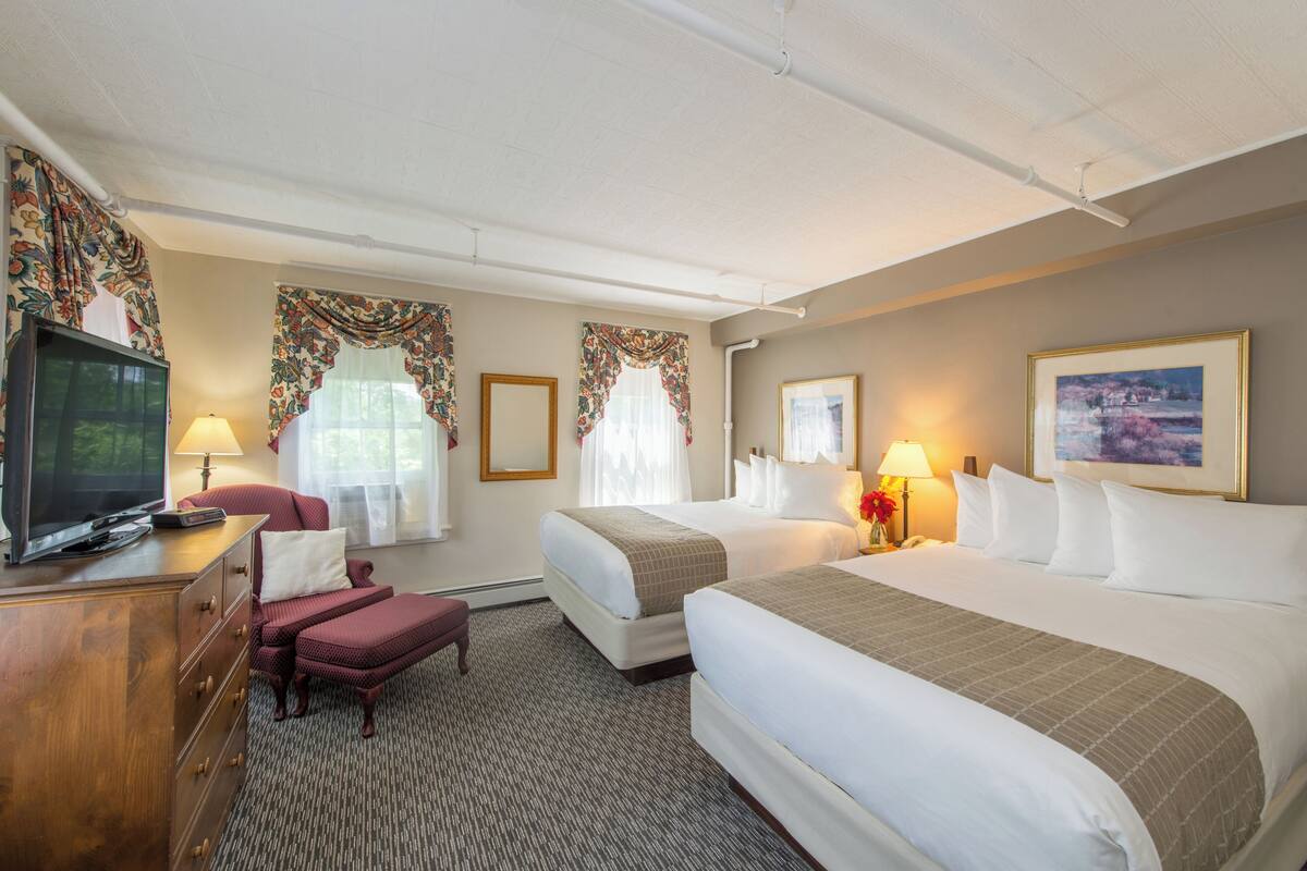 Quarto de hotel com duas camas, uma escrivaninha com televisão, uma cadeira e um abajur.