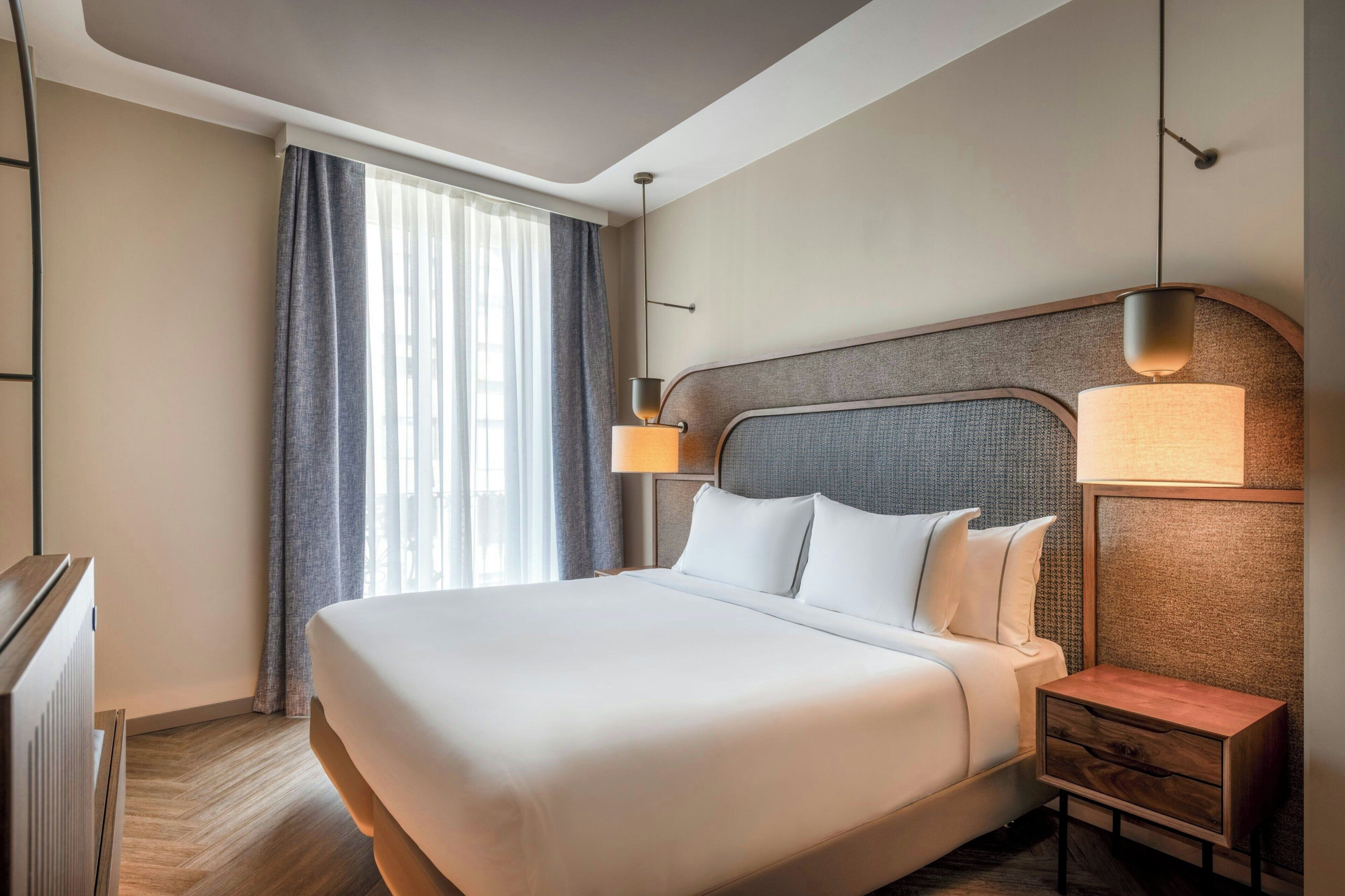 Quarto de hotel com uma cama grande, uma mesinha de cabeceira de madeira, uma luminária e vista para o exterior através de uma janela com cortinas.