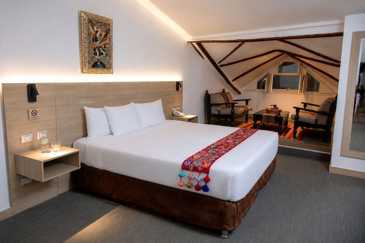 Quarto de hotel com uma cama grande, uma mesinha de cabeceira, uma cadeira, uma pequena mesa e um objeto decorativo na parede.