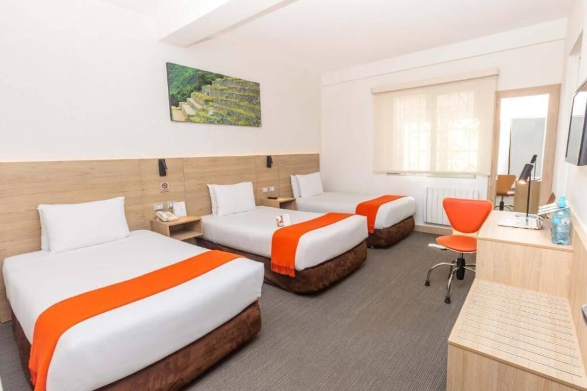 Quarto de hotel com três camas, uma escrivaninha, uma cadeira e uma janela com cortinas.