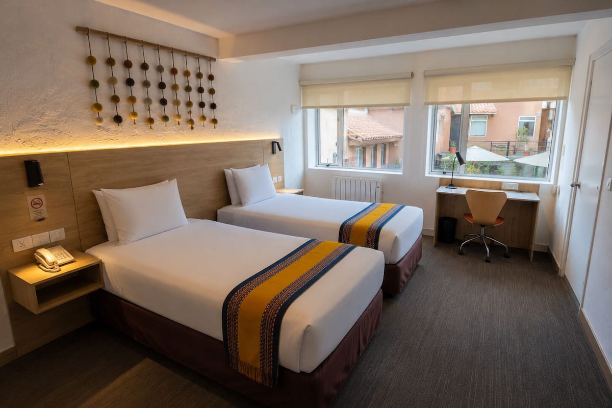 Quarto de hotel com duas camas, uma escrivaninha, uma cadeira, um telefone, uma luminária na parede e vista para o exterior.