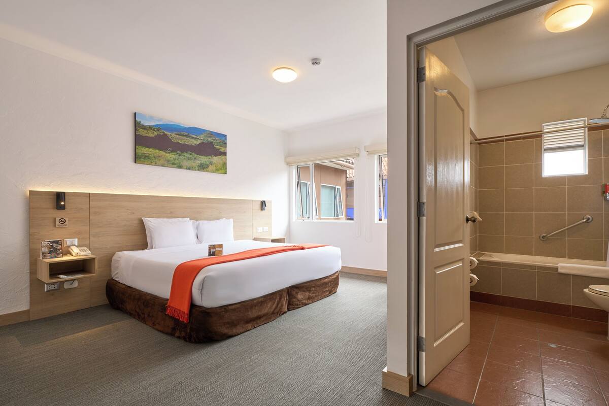 Quarto de hotel com cama, mesa de cabeceira, um quadro, banheiro com chuveiro e janela.